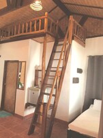 Quarto com escada de madeira para um mezanino, uma cama, cômoda pequena e espelho.
