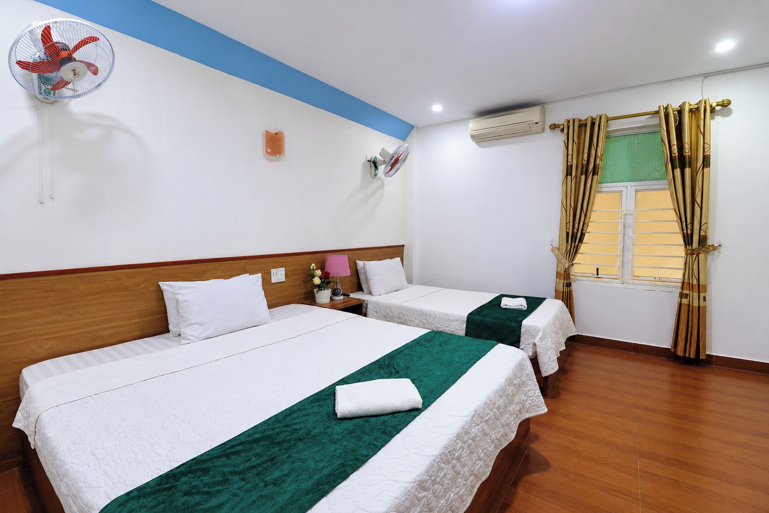 Quarto de hotel com duas camas, cabeceira de madeira, ar condicionado fixado na parede, janela com cortinas e um ventilador de parede.