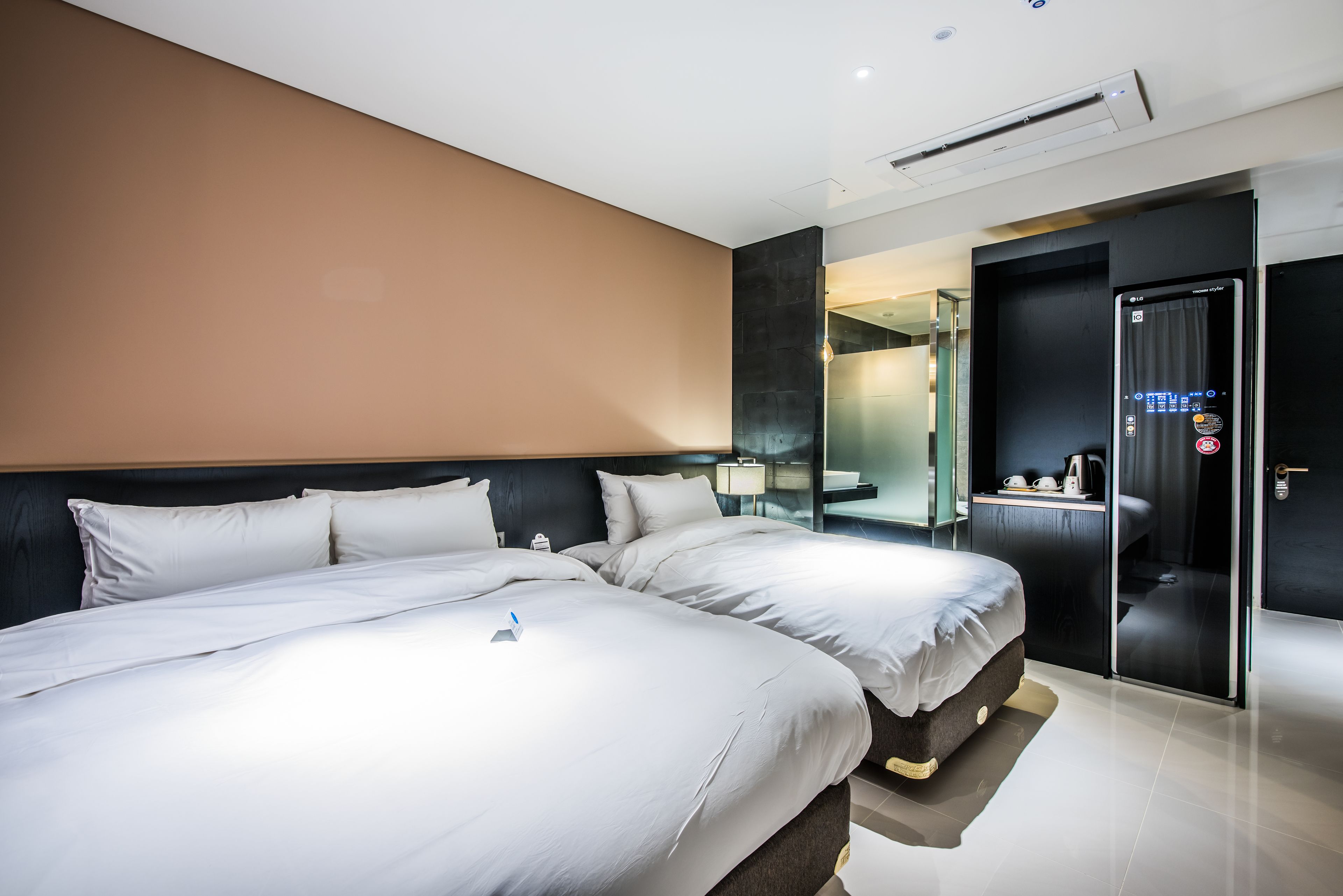 Quarto de hotel moderno com duas camas, um espelho grande, uma escrivaninha e um banheiro visível por uma porta de vidro.
