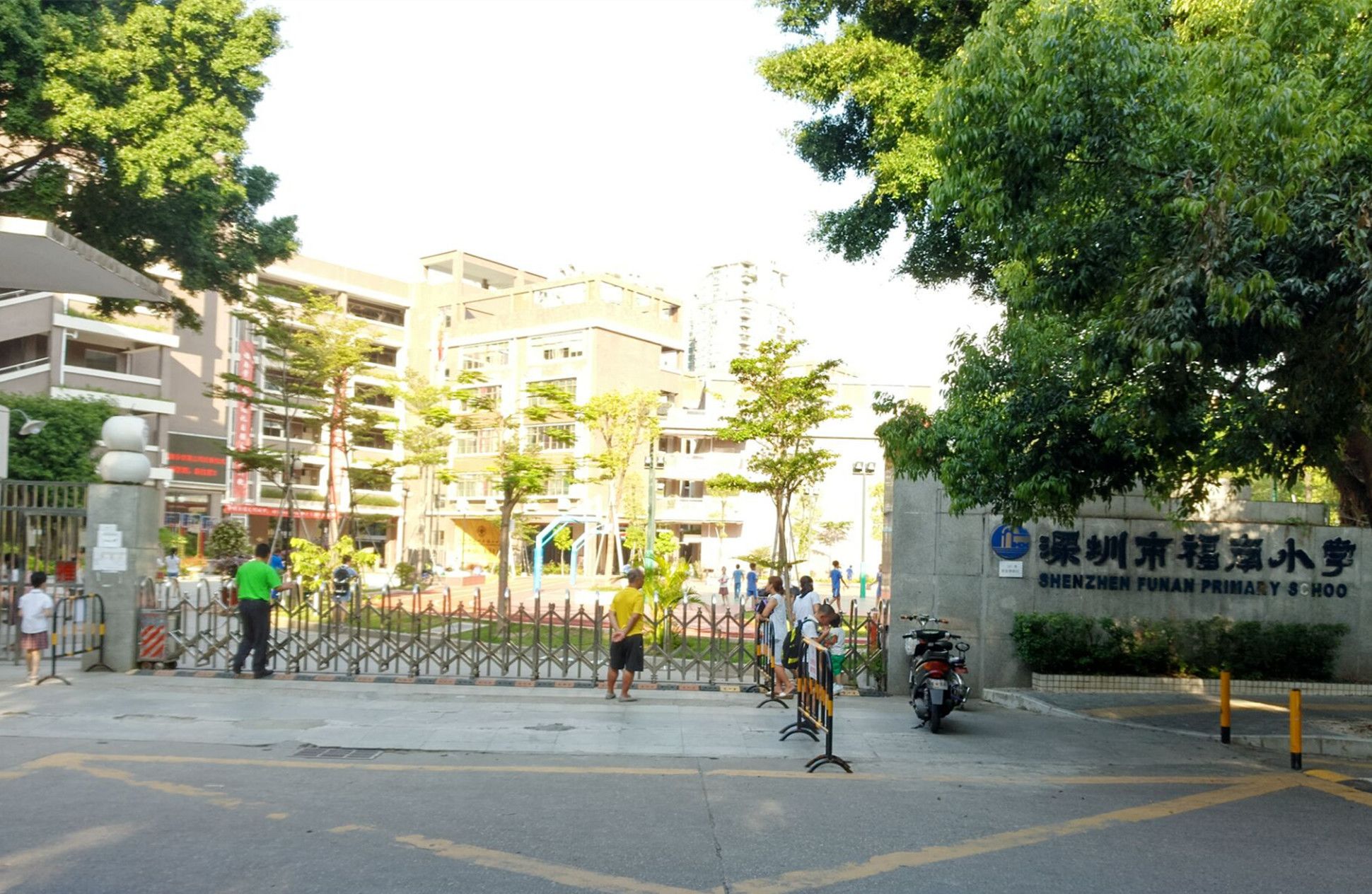 Entrada de escola primária com uma placa, uma motocicleta e pessoas caminhando.
