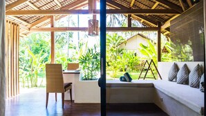 Um quarto com piso de madeira, uma cadeira de vime, um sofá com almofadas, uma mesa pequena e vista para o verde exuberante lá fora.