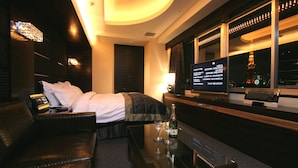 Quarto de hotel com cama, sofá, escrivaninha com computador e televisão exibindo uma lista de nomes.