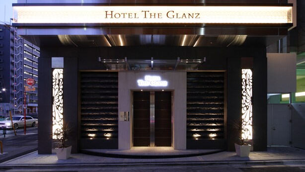 Hotel The Glanz com uma entrada moderna, grandes portas de vidro e uma placa iluminada de destaque.