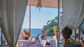 Uma área de refeições ao ar livre com toalhas de mesa xadrez, vista para o mar e um vaso de planta.