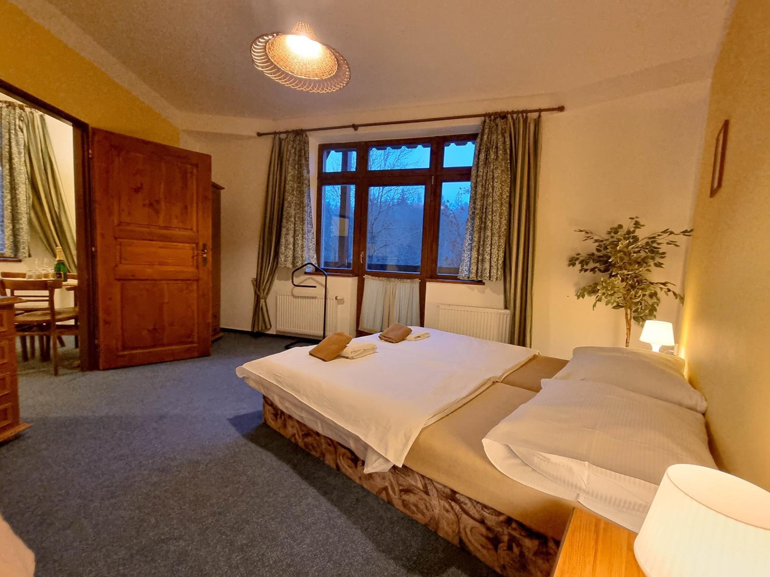 Quarto com cama, escrivaninha, cadeira, janela com cortinas, radiador e planta.