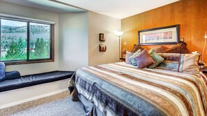 Quarto com cama grande, um banquinho aos pés da cama, janela com vista para árvores e um quadro na parede.