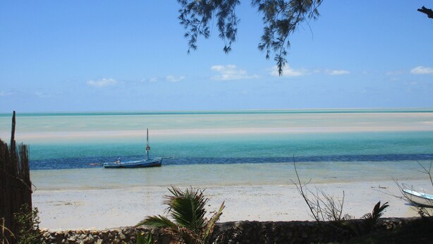 Uma praia com águas turquesa cristalinas, um barco na areia e um muro de pedra em primeiro plano.