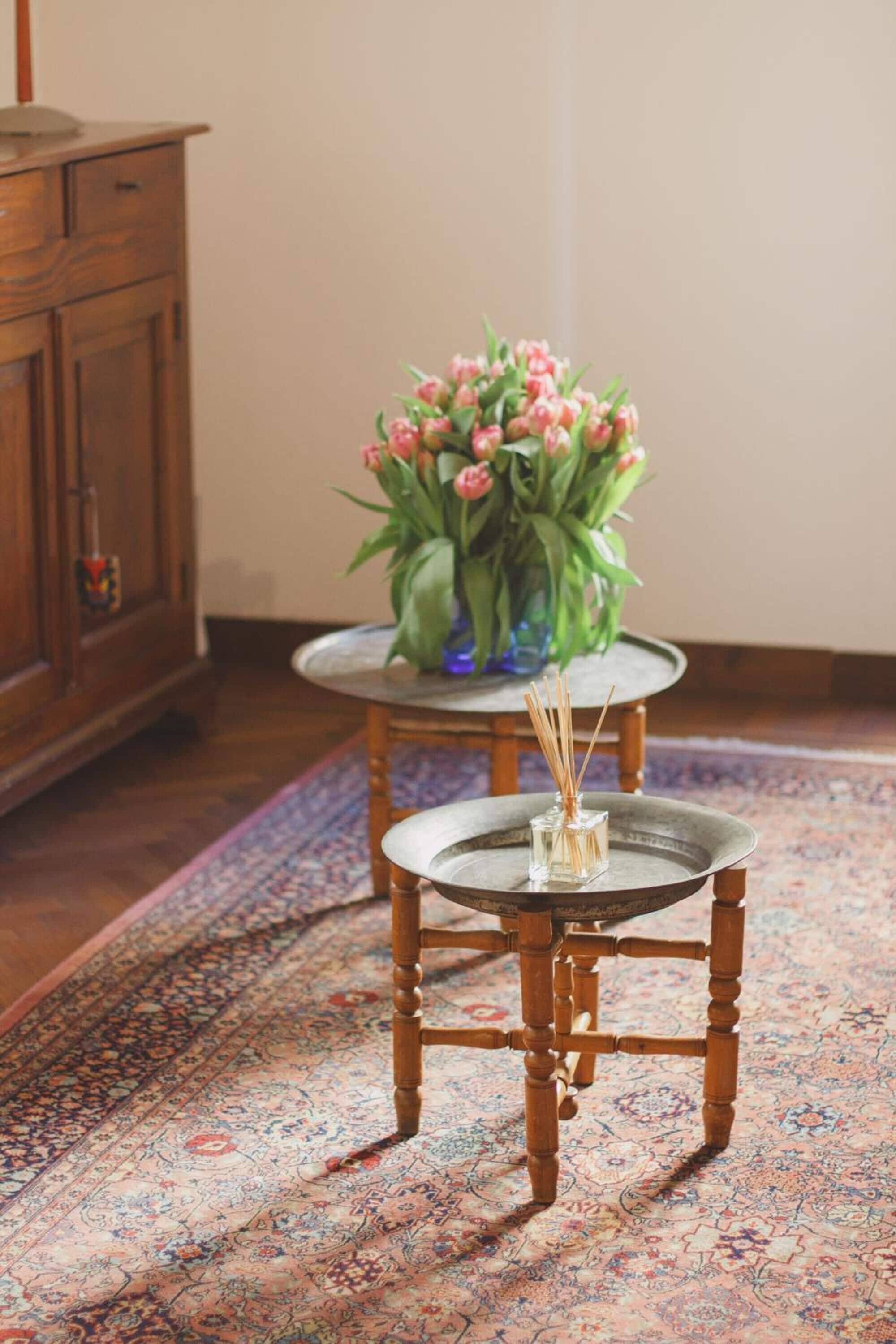 Uma cadeira de madeira com uma mesinha de bandeja, um vaso com flores cor-de-rosa e um armário de madeira em um quarto com carpete estampado.