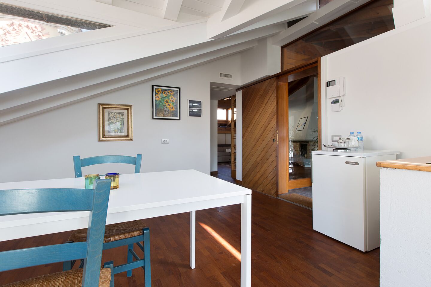 Uma cozinha compacta com uma mesa branca e cadeiras azuis, chão de madeira e vista para a área de estar vizinha.