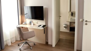 Quarto de hotel com escrivaninha, cadeira, televisão e banheiro visível por uma porta aberta.