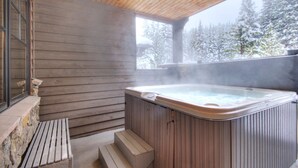 Um deck de madeira com hidromassagem, degraus de pedra e vista para árvores cobertas de neve.