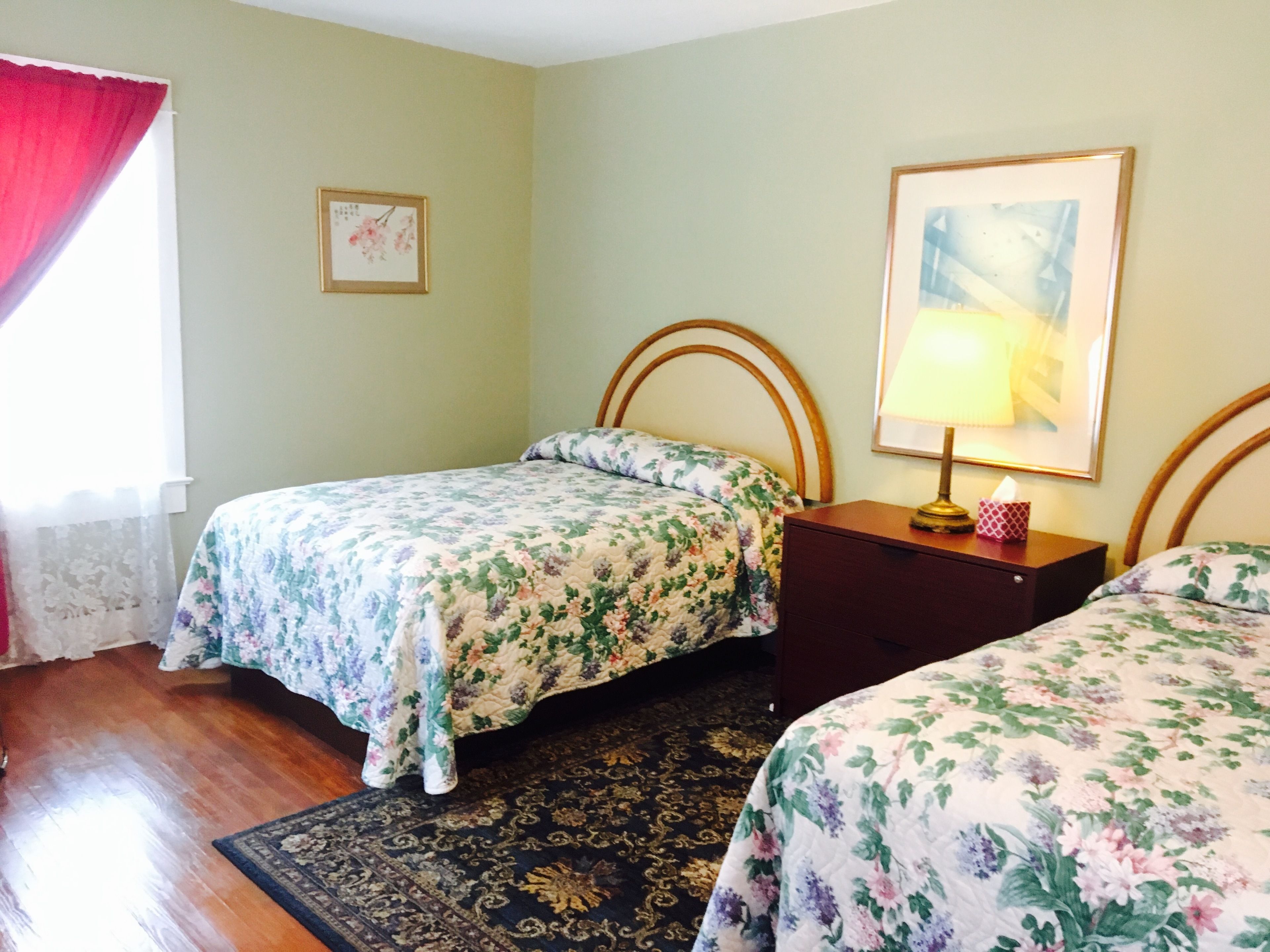 Quarto com duas camas de solteiro, mesa de cabeceira com abajur, quadro emoldurado na parede e janela com cortina vermelha.