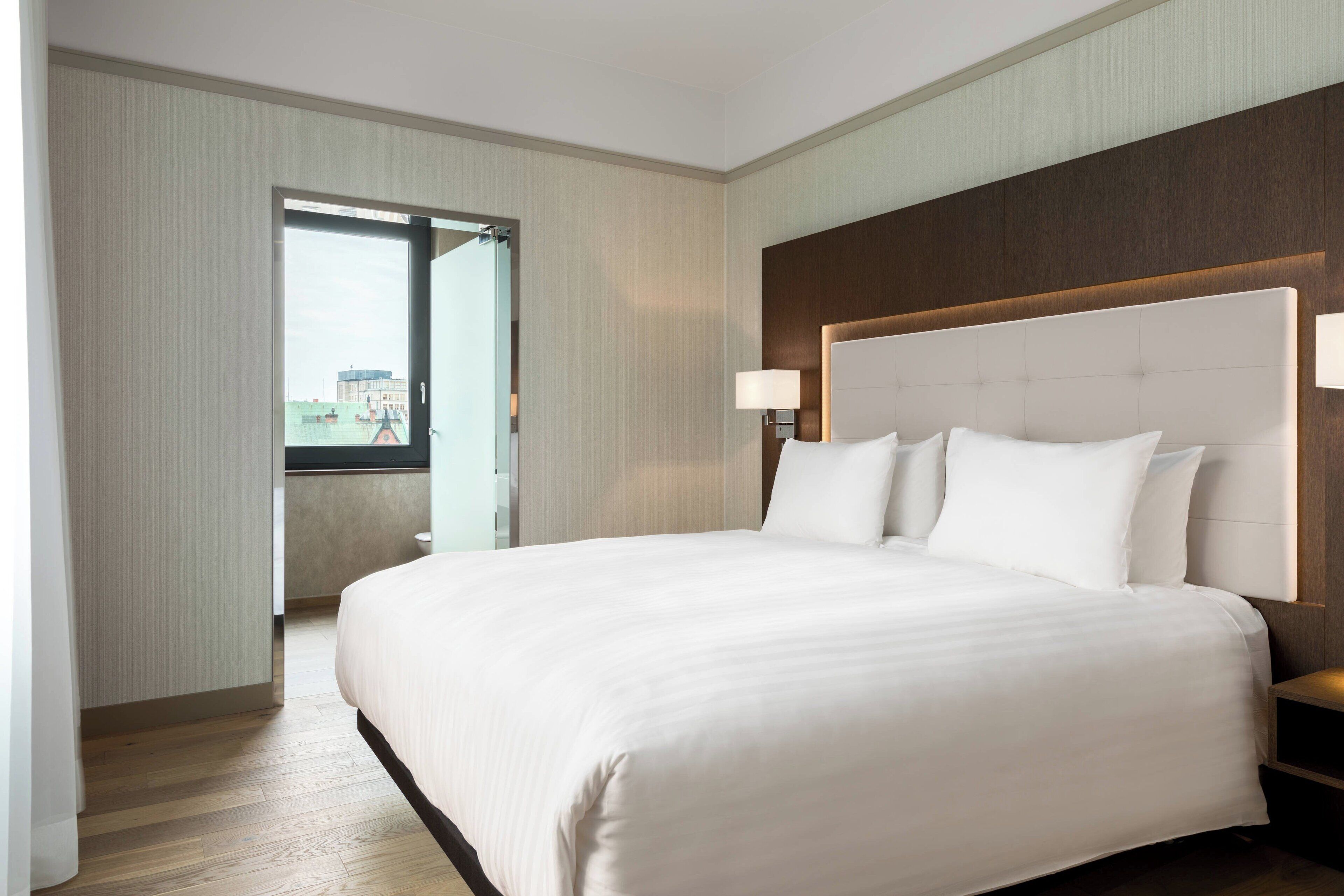 Quarto de hotel com uma cama grande, cabeceira de madeira, mesa de cabeceira e janela com vista para edifícios.