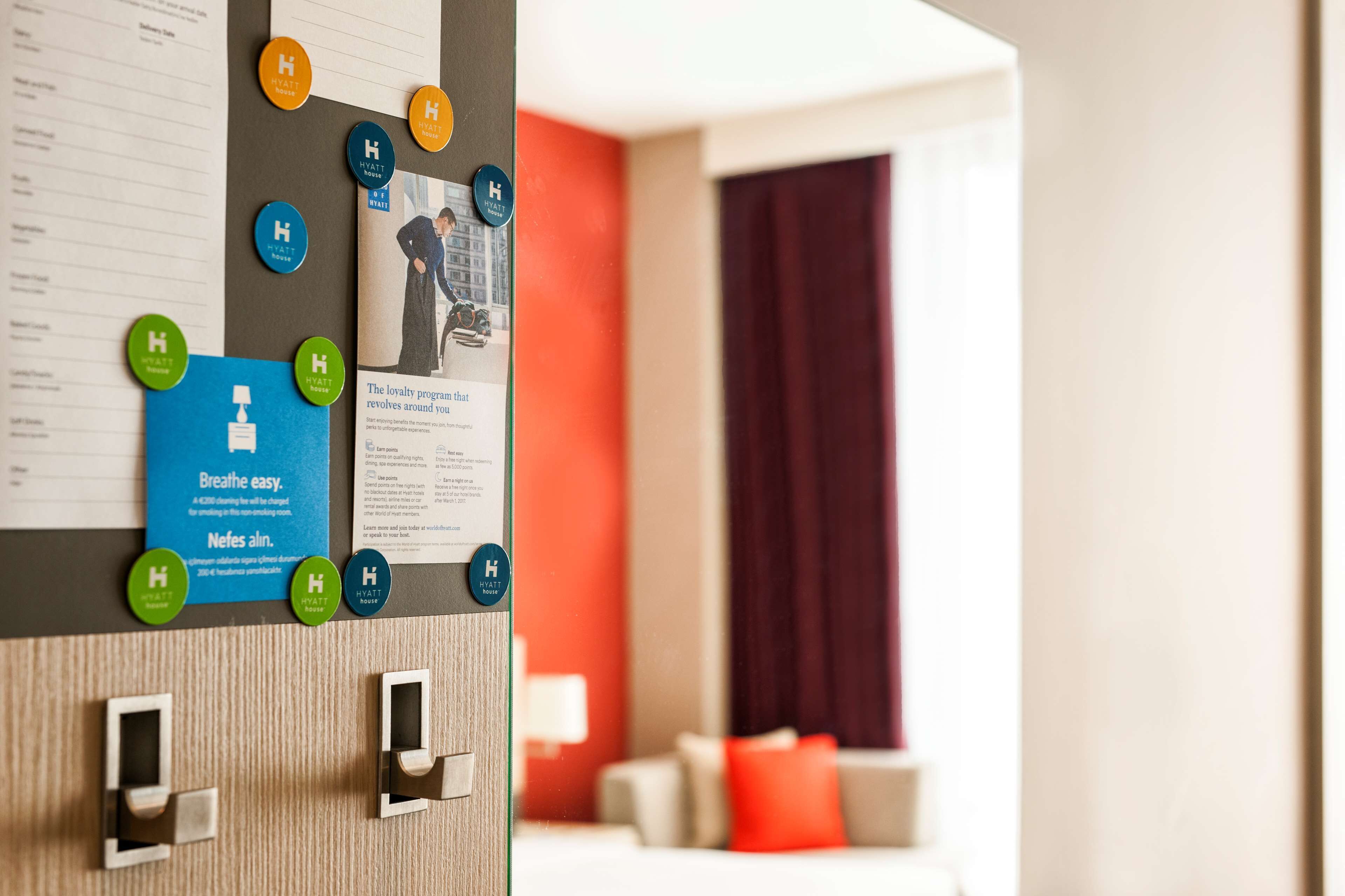 Quarto de hotel com um mural exibindo um cartaz de programa de fidelidade, um painel de controle e uma cortina vermelha ao fundo.