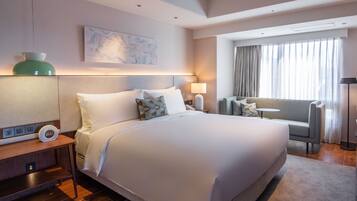Quarto de hotel moderno com uma cama grande, mesas de cabeceira, uma escrivaninha, uma cadeira e uma janela ampla com cortinas.