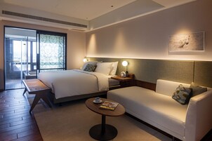 Quarto de hotel com uma cama, um sofá, uma mesinha de centro com uma revista e uma mesa de cabeceira com abajur.