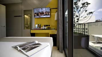 Quarto de hotel moderno com uma cama, televisão na parede, varanda com vista para a cidade e pequena área de cozinha.