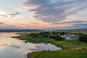 Uma paisagem tranquila à beira do lago, com um pequeno edifício branco próximo à água, cercado por vegetação e algumas outras construções.