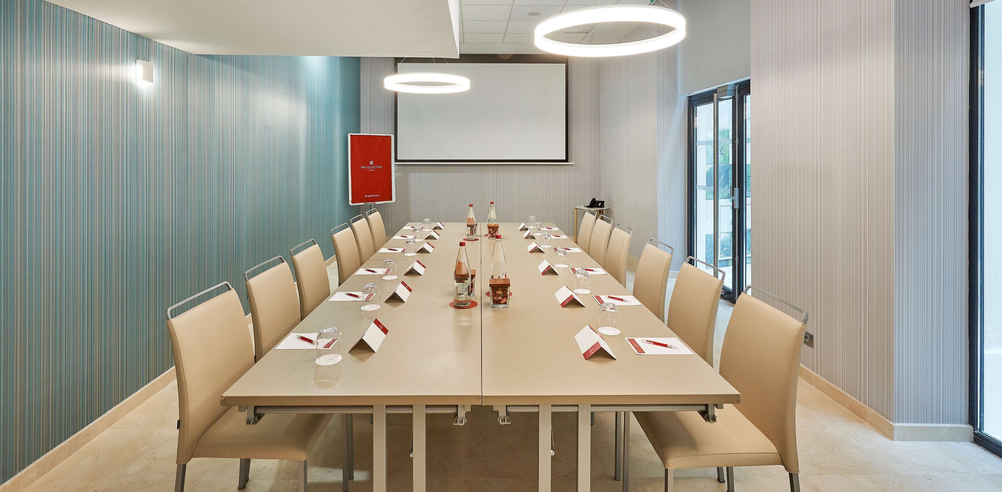 Sala de conferências com uma mesa comprida, cadeiras bege, cartões de nome e uma tela de projeção.