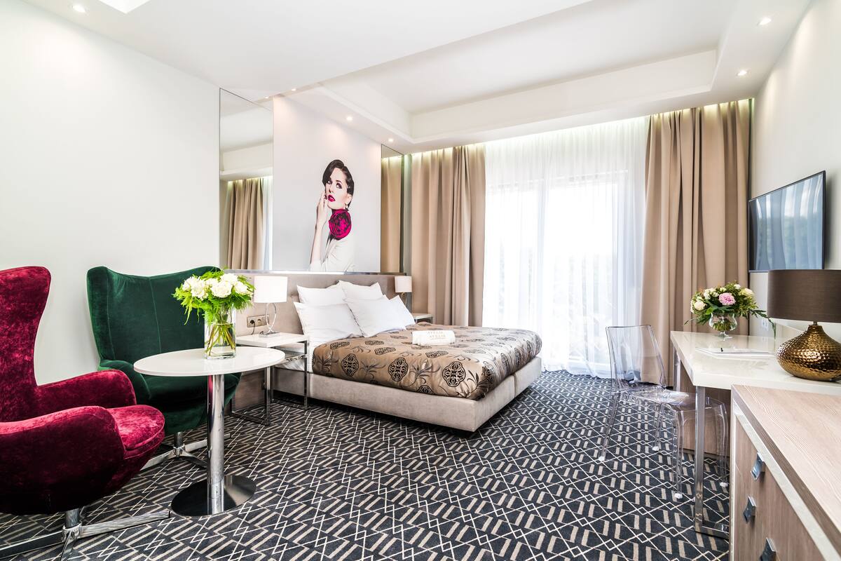 Quarto de hotel com cama, uma cadeira vermelha, uma mesa redonda pequena com um vaso, uma TV e um espelho.