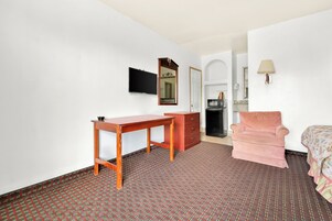 Quarto de hotel com uma escrivaninha de madeira, penteadeira, poltrona rosa, televisão, espelho e abajur.