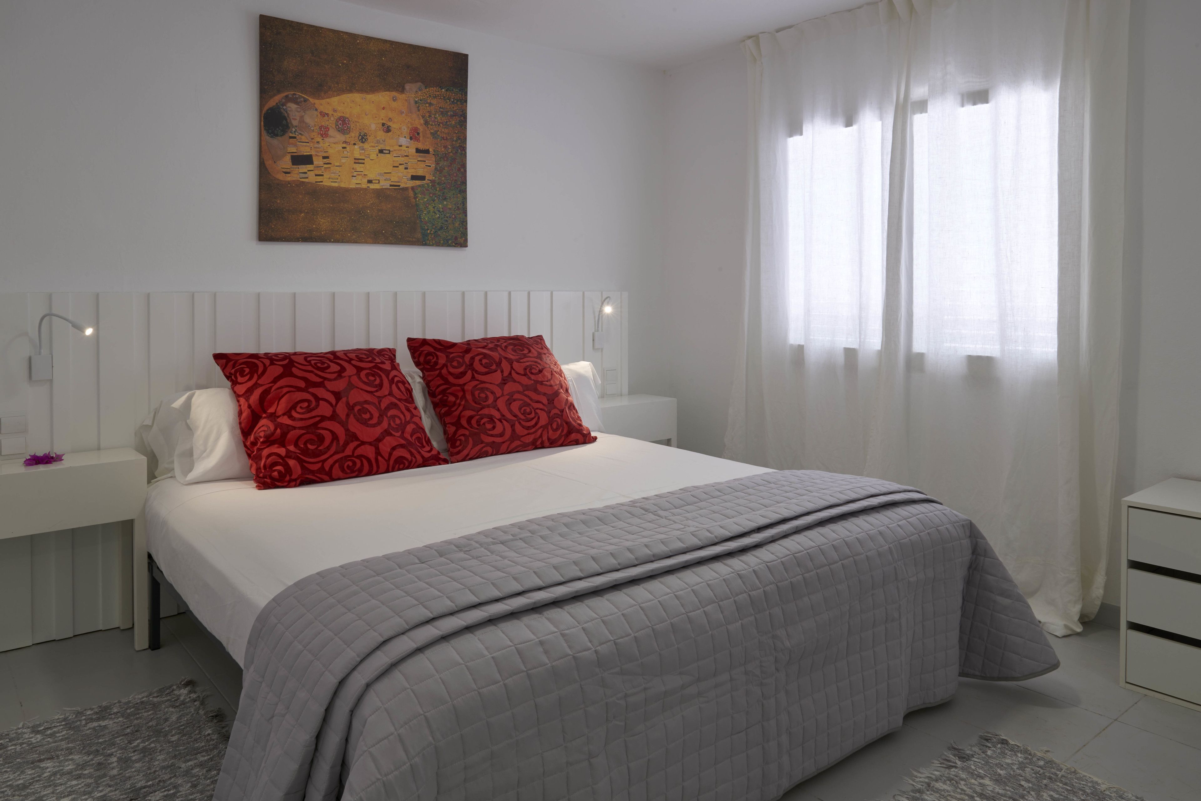 Quarto com cama, mesas de cabeceira brancas, um quadro na parede e cortinas leves.
