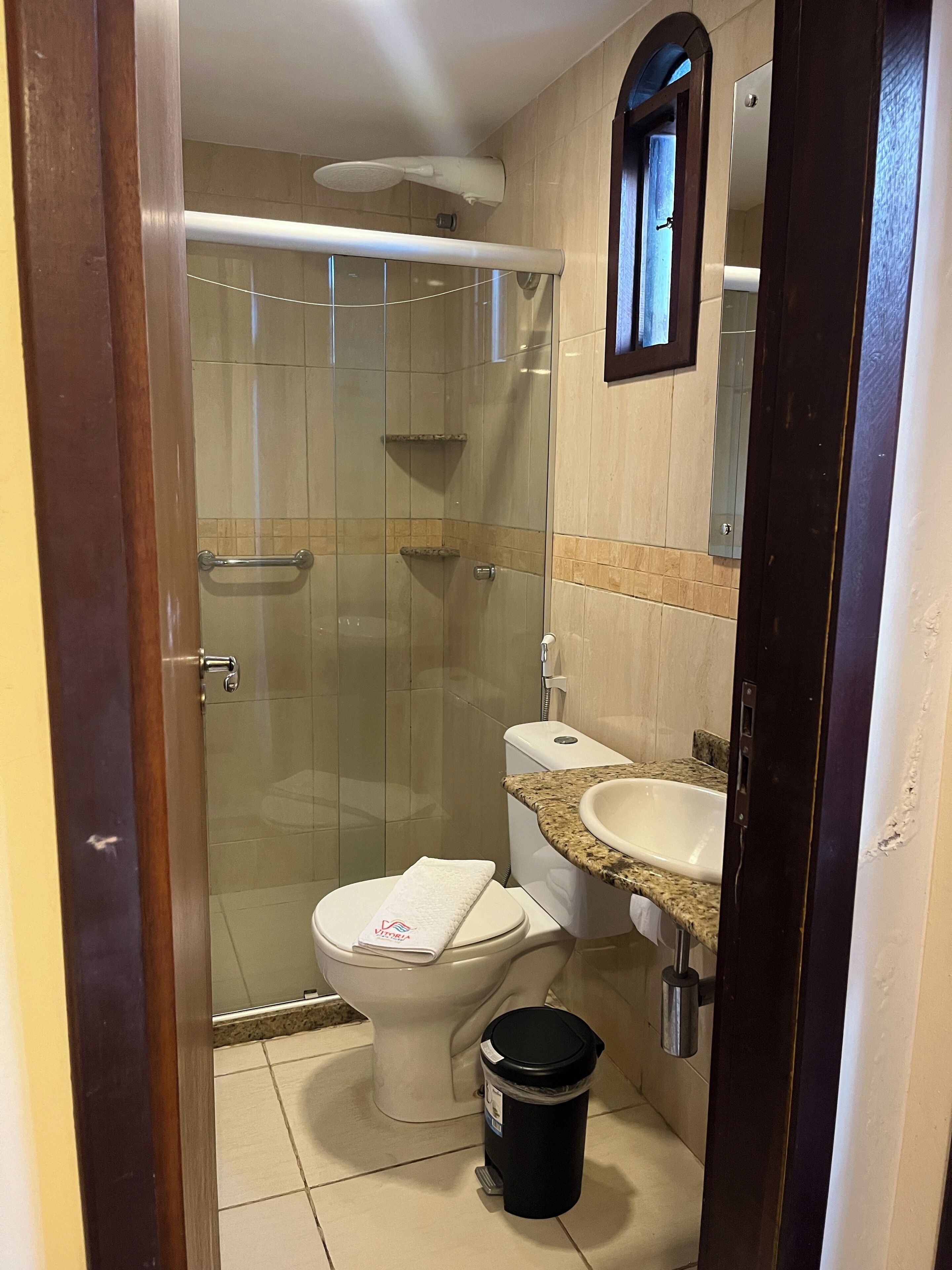 Casa de banho com chuveiro envidraçado, sanita, lavatório e um contentor de lixo.