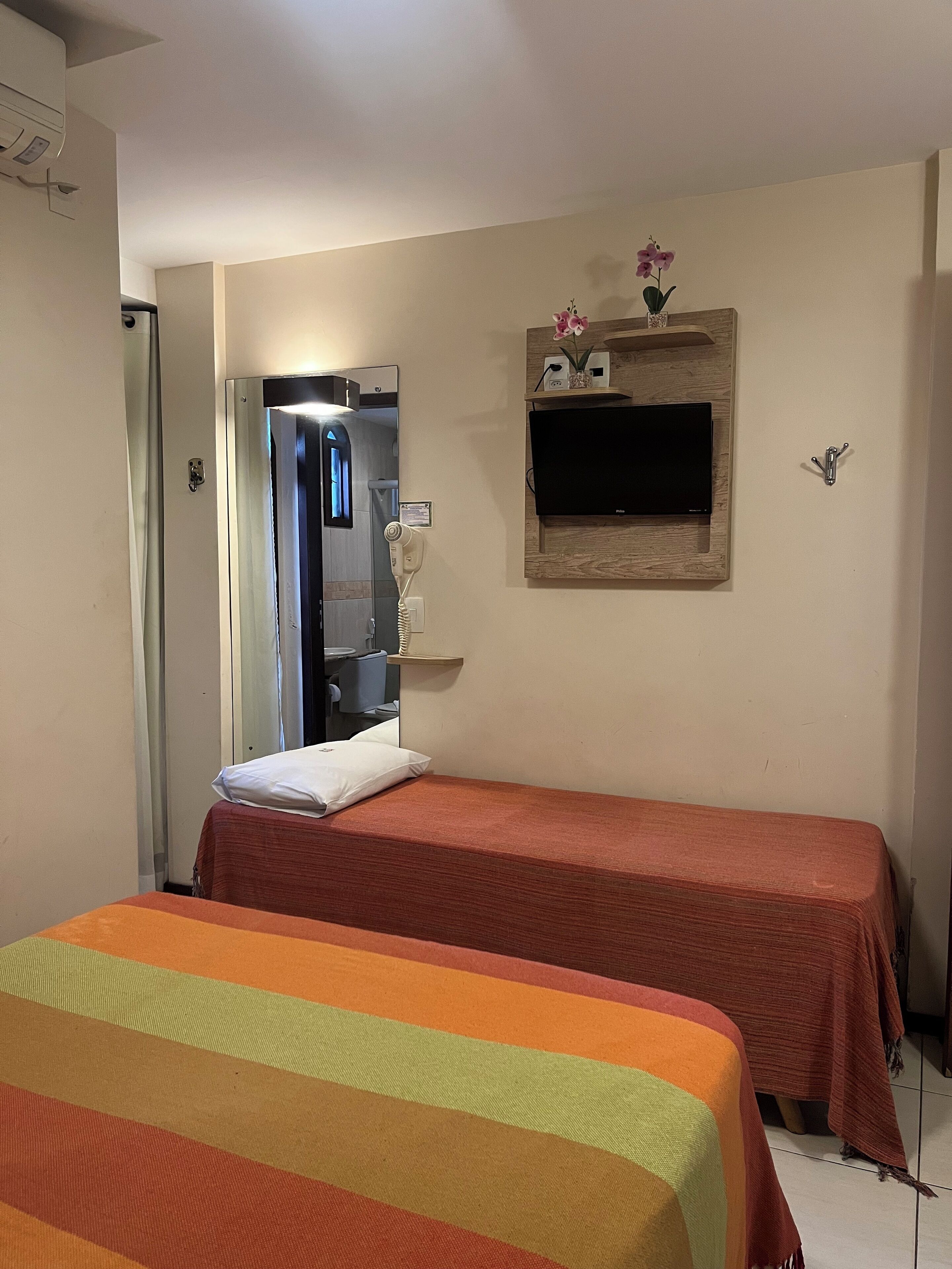 Quarto de hotel com duas camas de solteiro, televisão fixada na parede, um espelho e uma pequena estante com objetos decorativos.