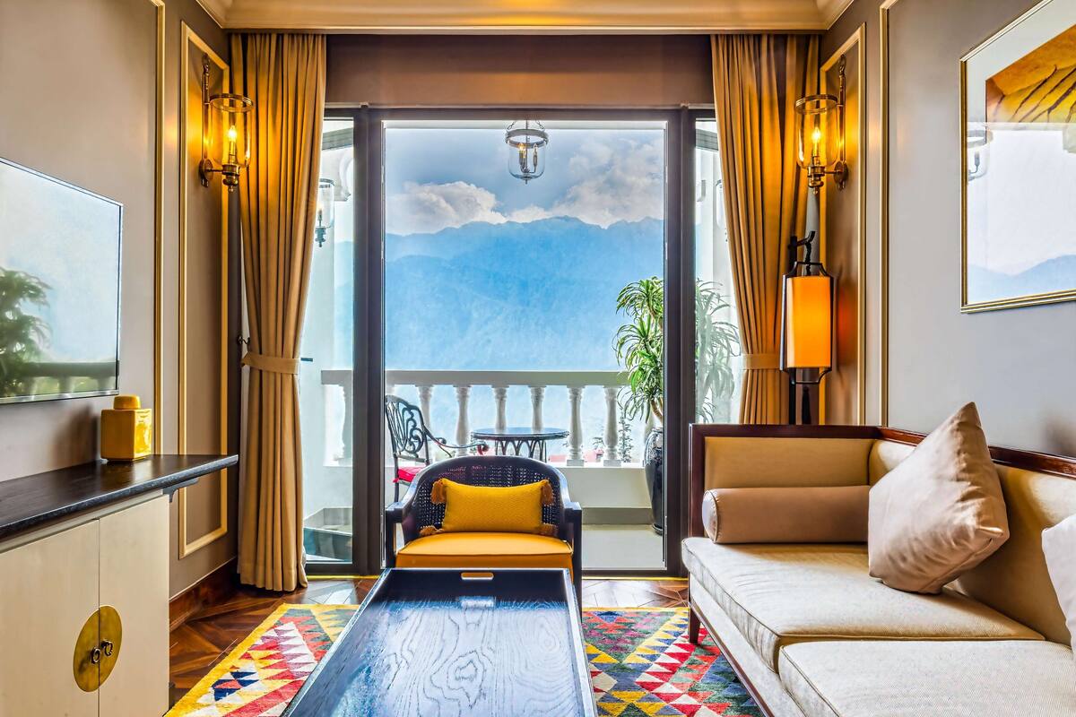 Quarto de hotel com varanda, sofá, cadeira, mesa de centro e vista para montanhas.