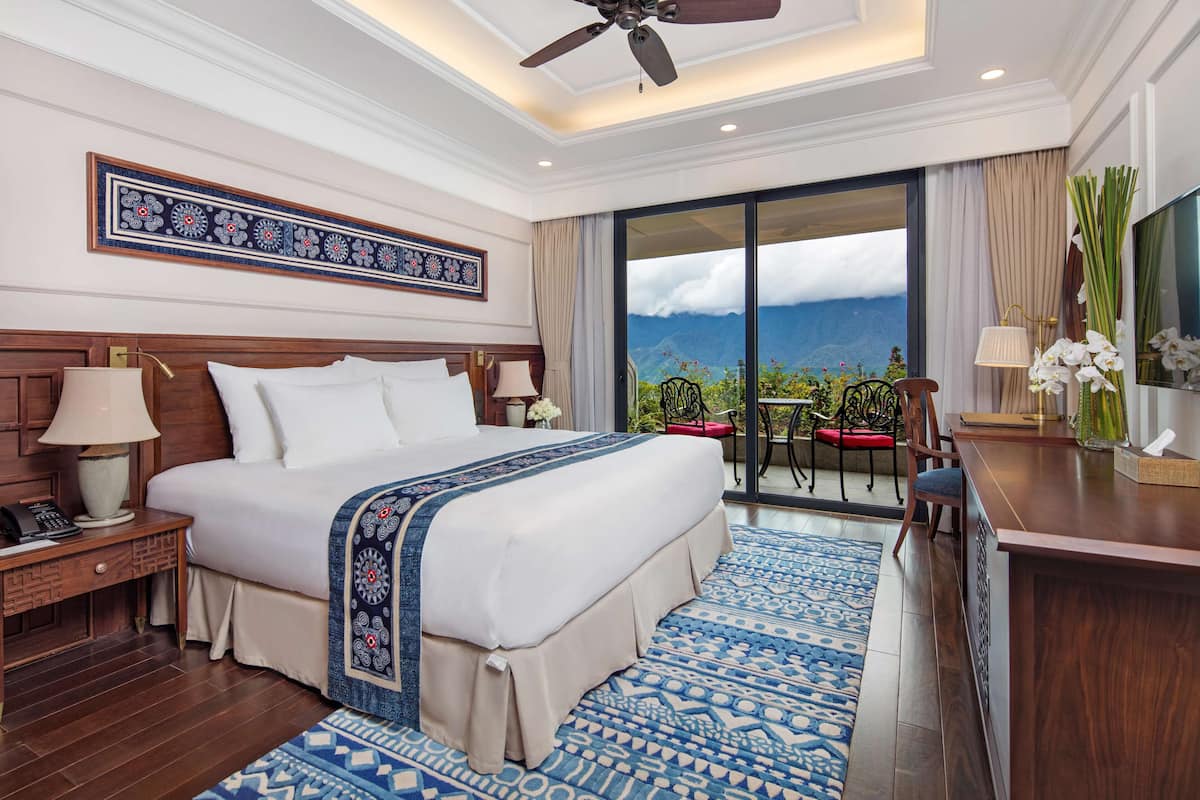 Quarto de hotel com uma cama grande, uma escrivaninha, uma cadeira, televisão e varanda com vista para as montanhas.