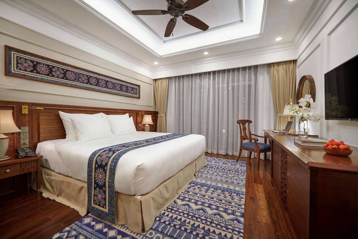Quarto de hotel com uma cama grande, uma escrivaninha, uma cadeira, um ventilador de teto e um detalhe decorativo na parede.