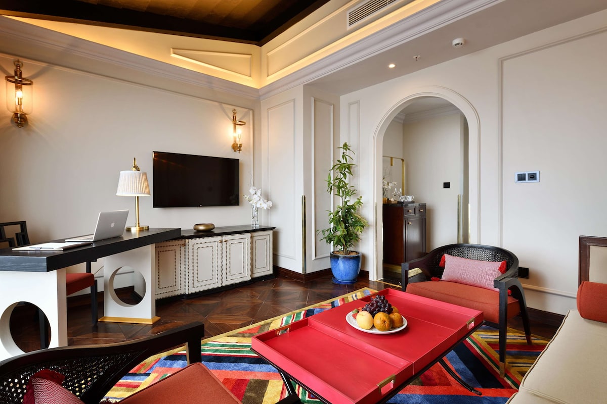 Quarto de hotel moderno com área de refeições, TV de tela plana, laptop, planta em vaso e tapete colorido.