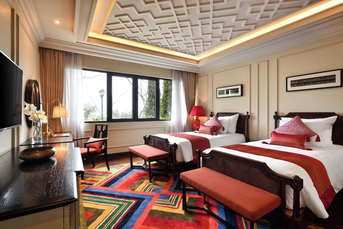 Quarto de hotel com duas camas, uma escrivaninha com televisão e um tapete colorido.