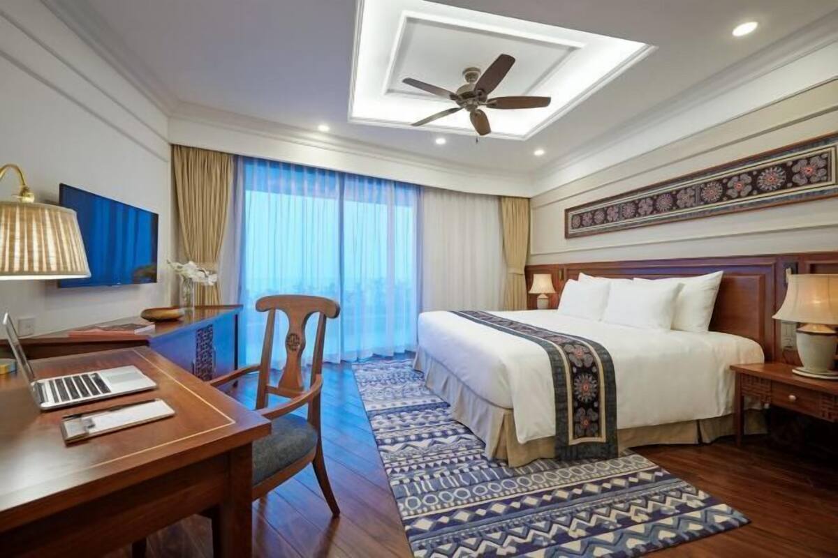Quarto de hotel com cama, escrivaninha, cadeira, ventoinha no teto e painel decorativo na parede.