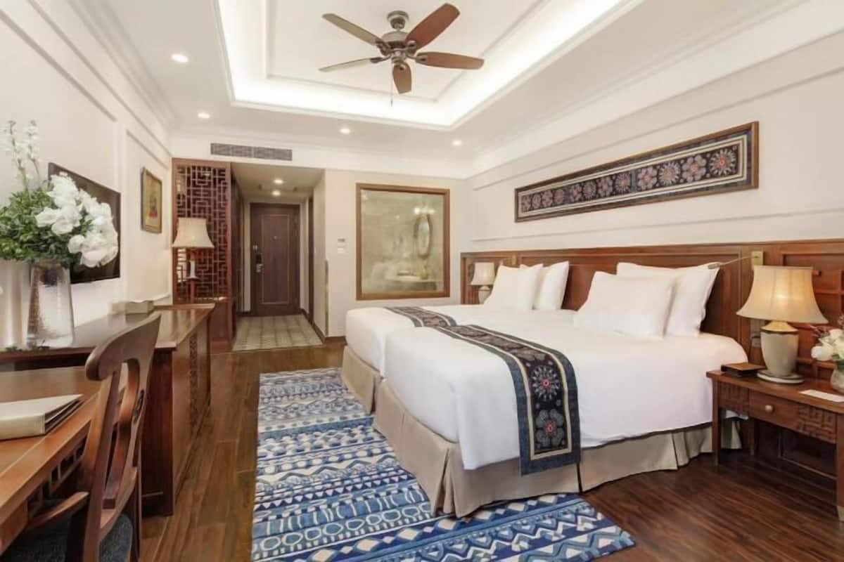 Quarto de hotel com cama, escrivaninha, cadeira, ventoinha no teto e tapete estampado.