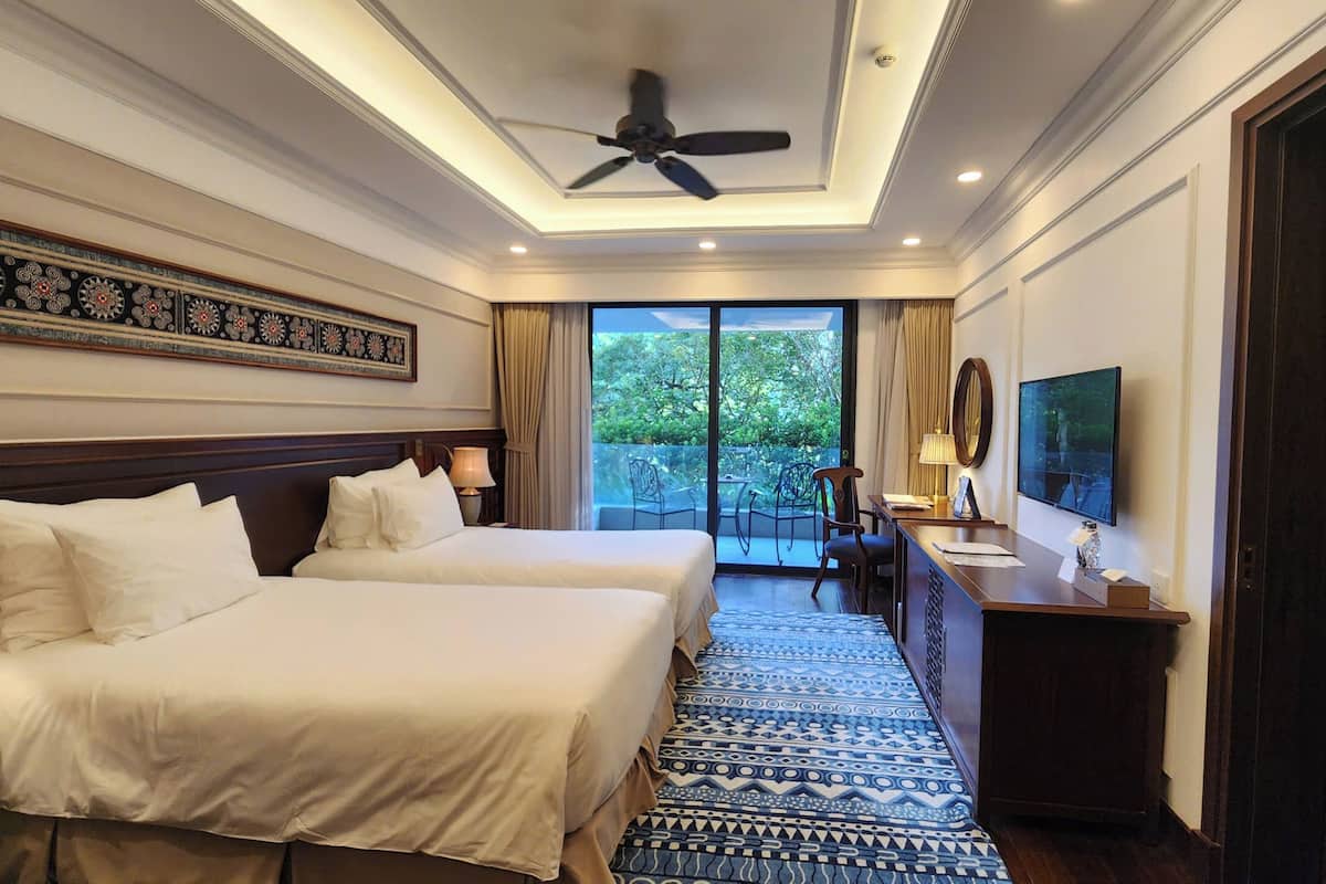 Quarto de hotel com duas camas, uma escrivaninha com cadeira, televisão e varanda com vista para o verde.
