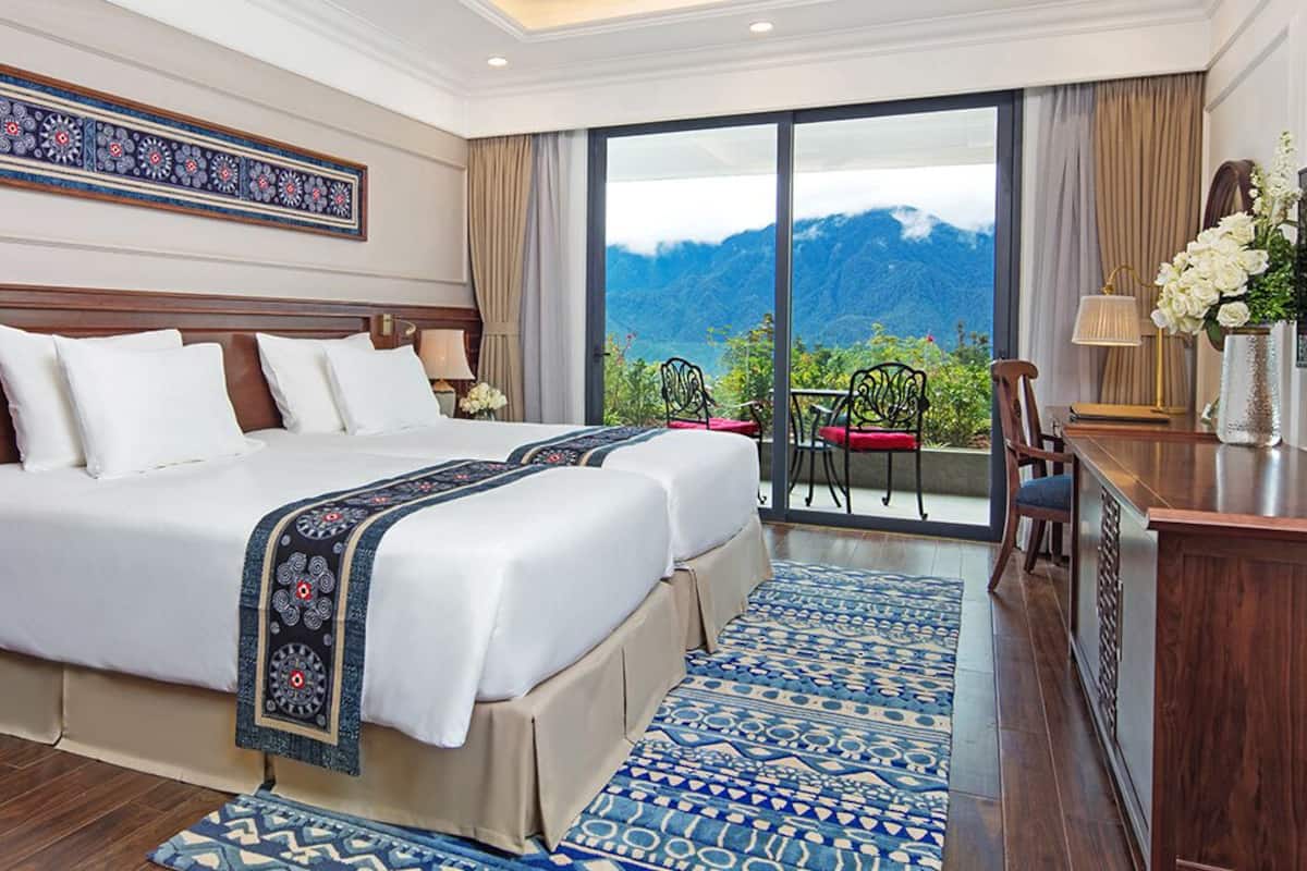 Quarto de hotel com uma cama grande, uma mesa de trabalho, televisão e varanda com vista para as montanhas.