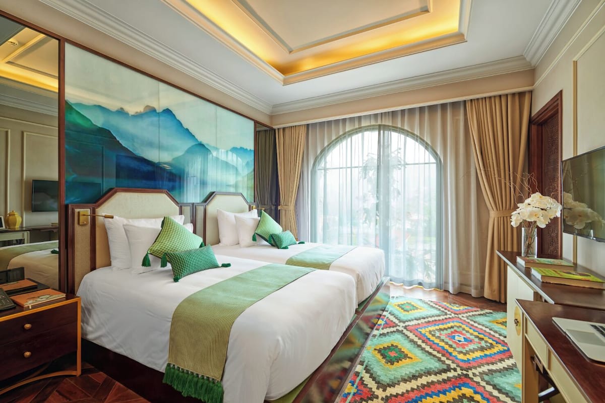 Quarto de hotel com duas camas, um quadro grande, uma escrivaninha com um computador e uma janela ampla com cortinas.