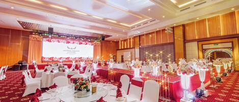 Uma sala de banquetes com mesas redondas preparadas para refeições, cadeiras brancas e um palco com fundo vermelho.