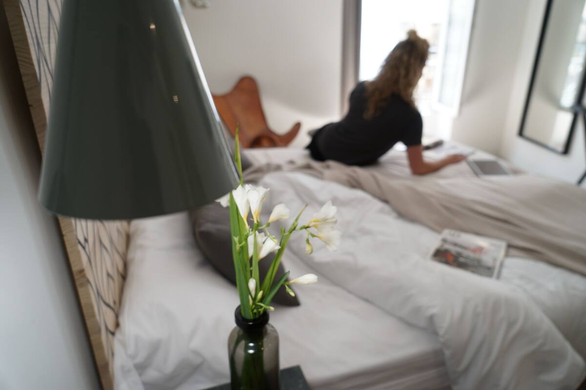 Uma cama com roupa de cama branca, um vaso com flores brancas e um abajur.