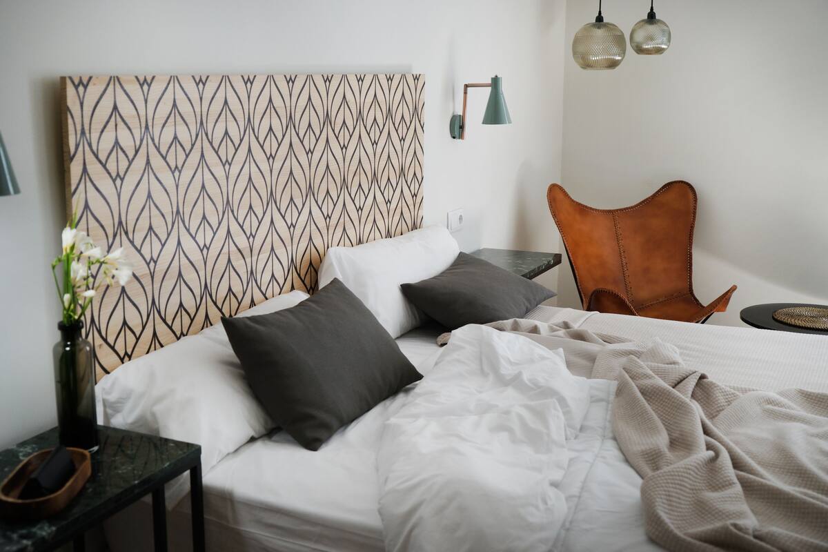 Cama bem arrumada com almofadas, um vaso com flores e uma poltrona de couro marrom em um quarto moderno com cabeceira estampada e luminárias pendentes.