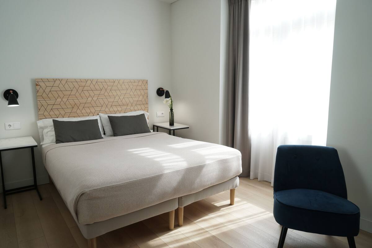Quarto moderno com cama grande, mesas de cabeceira, uma cadeira e cabeceira com padrão geométrico.