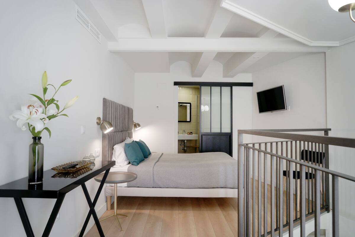 Quarto moderno com cama, mesa de cabeceira, abajur e vista para um banheiro.