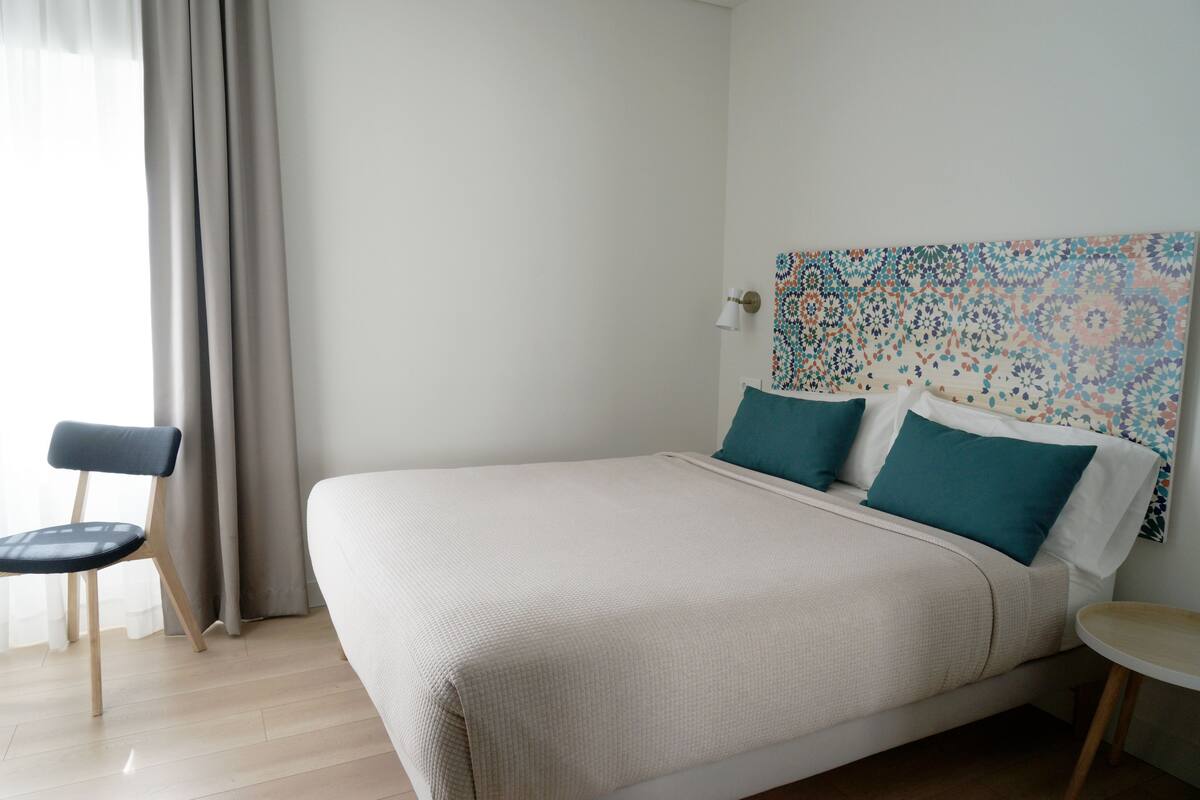 Quarto de hotel moderno com uma cama grande, uma mesa pequena, uma cadeira e um objeto decorativo na parede.