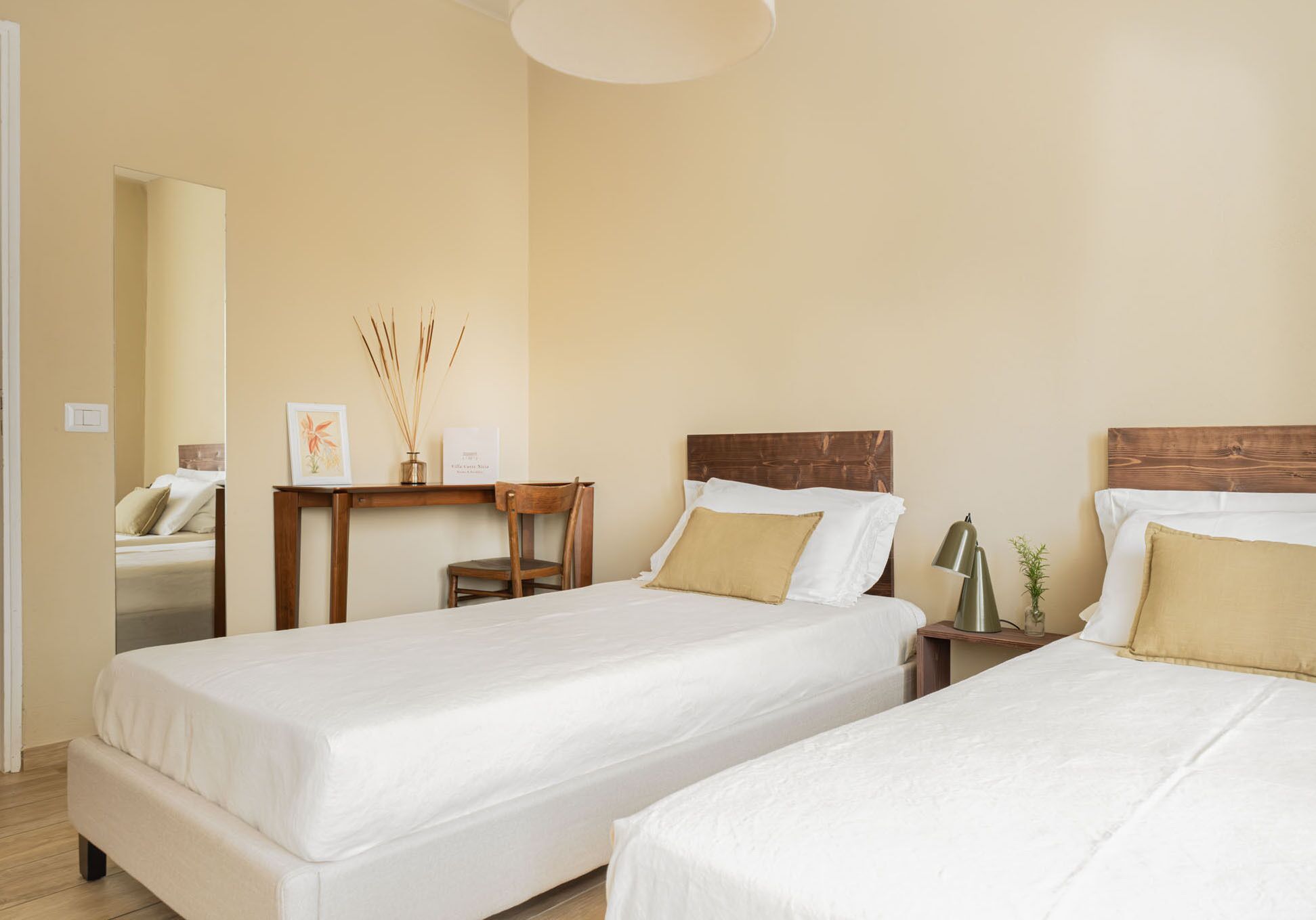 Quarto de hotel com duas camas, uma mesa pequena com cadeira, mesa de cabeceira com abajur e um espelho.