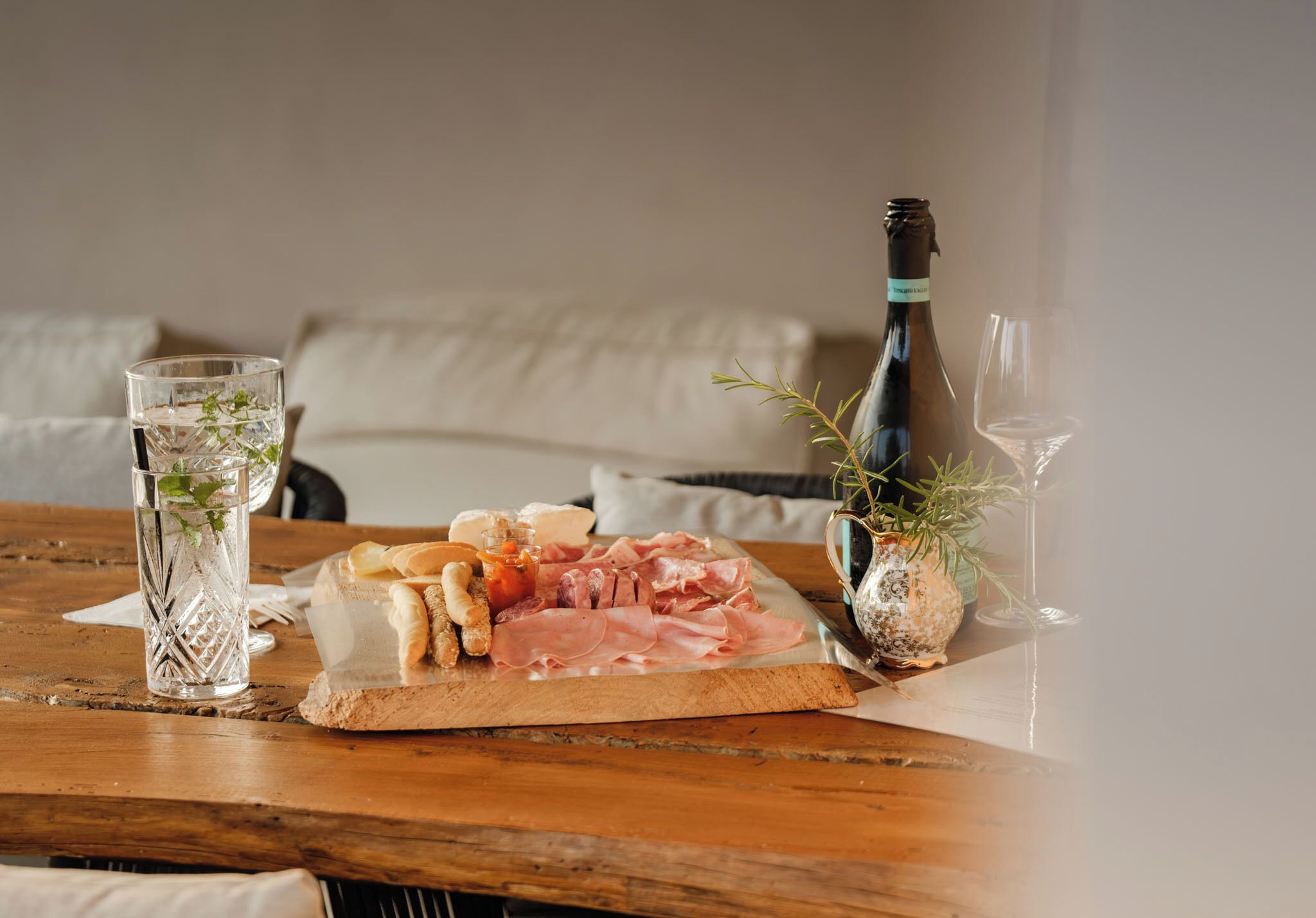Uma mesa de madeira com uma tábua de frios, uma garrafa de vinho, um copo de água e um vaso decorativo com uma planta.