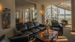 Uma sala de estar moderna com um sofá de couro preto, uma mesa de centro de madeira e um espelho grande que reflete o ambiente. Janelas amplas revelam a vista de um corpo d'água.