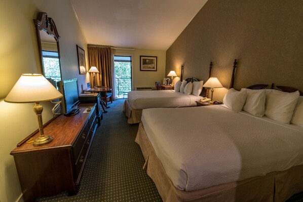 Quarto de hotel com duas camas, uma escrivaninha com luminária, uma cadeira e uma janela com cortinas.