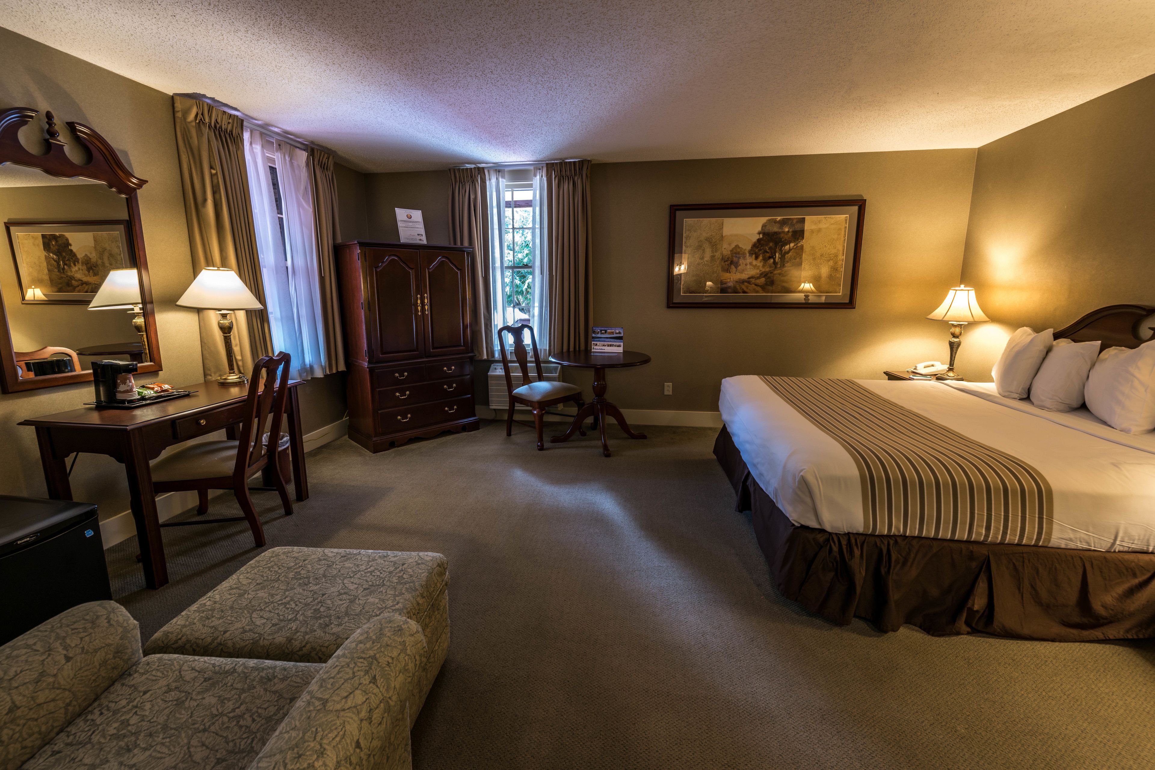 Quarto de hotel com cama, escrivaninha, cadeira, cômoda e janela com cortinas.