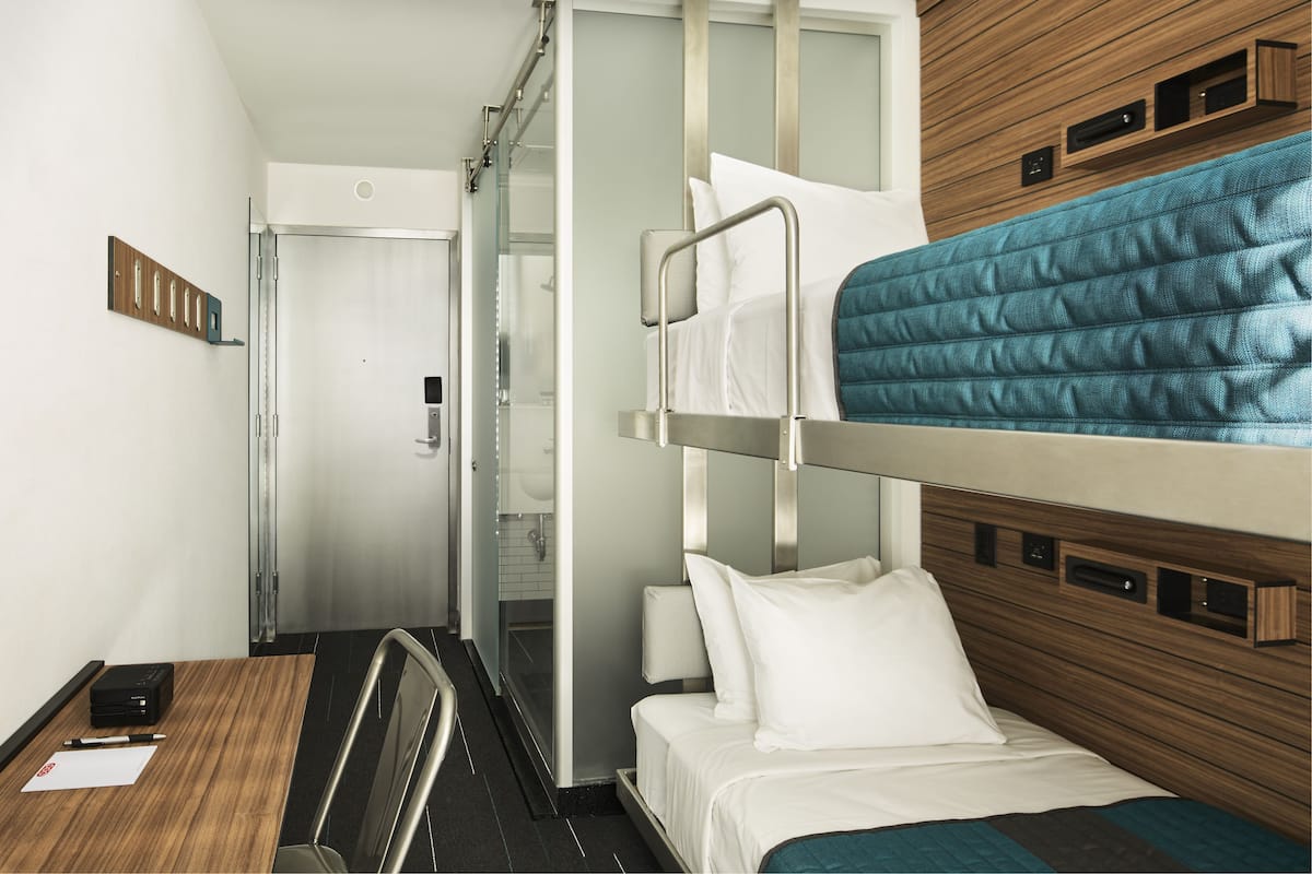 Quarto de hotel compacto com beliche, uma escrivaninha com cadeira e um banheiro pequeno visível através de uma porta de vidro.